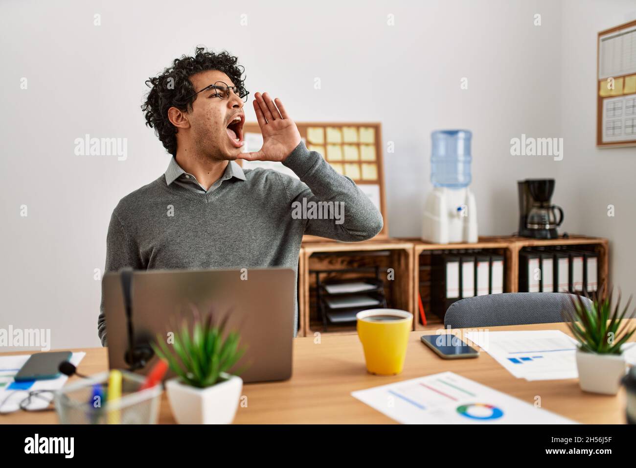 Young hispanic man wearing business style sitting on desk at office ...