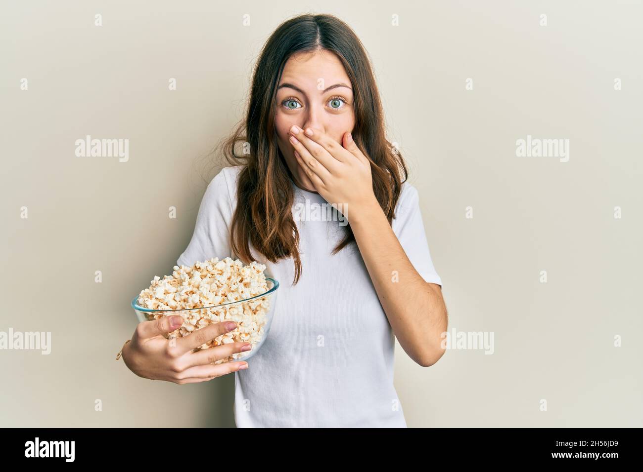 Young brunette woman eating popcorn covering mouth with hand, shocked ...