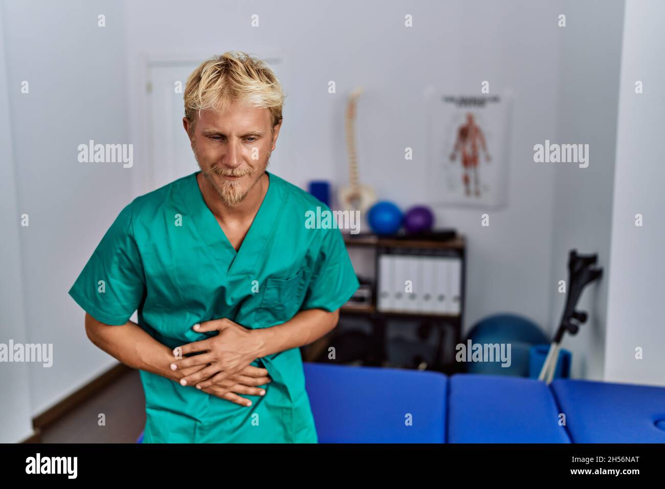 Young blond man wearing physiotherapist uniform standing at clinic with ...