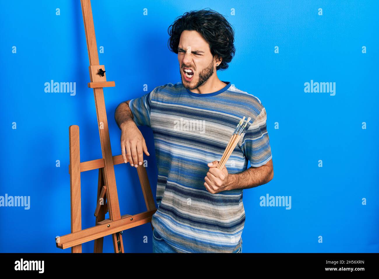 Handsome hispanic man standing by empty easel stand holding brushes ...