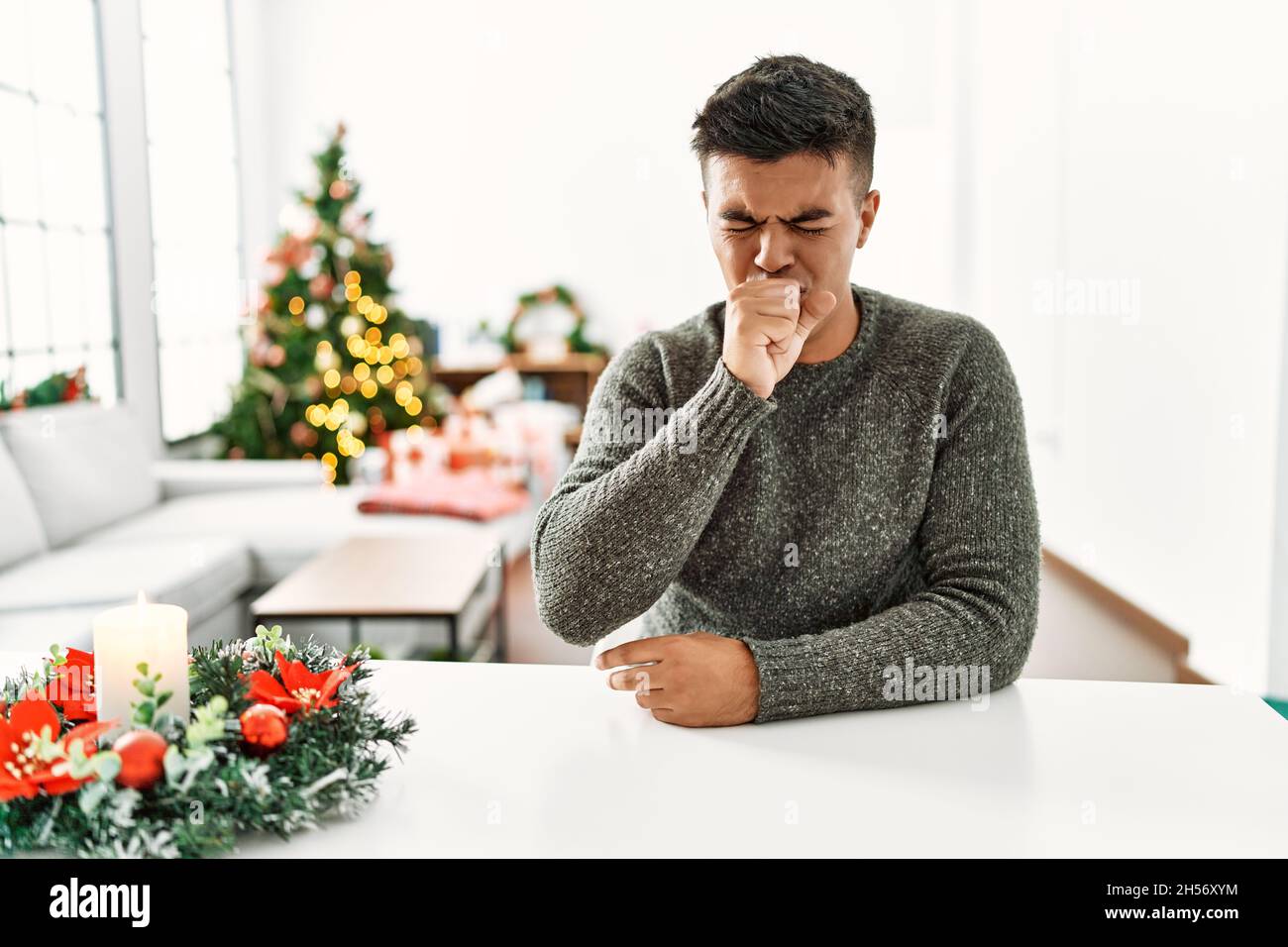 Young hispanic man sitting on the table by christmas tree feeling ...