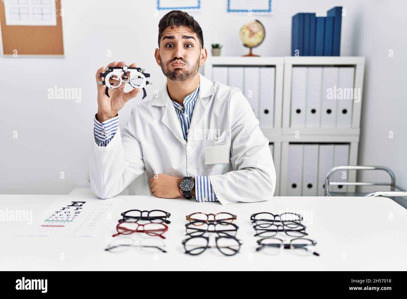 Young optician man holding optometry glasses puffing cheeks with funny ...