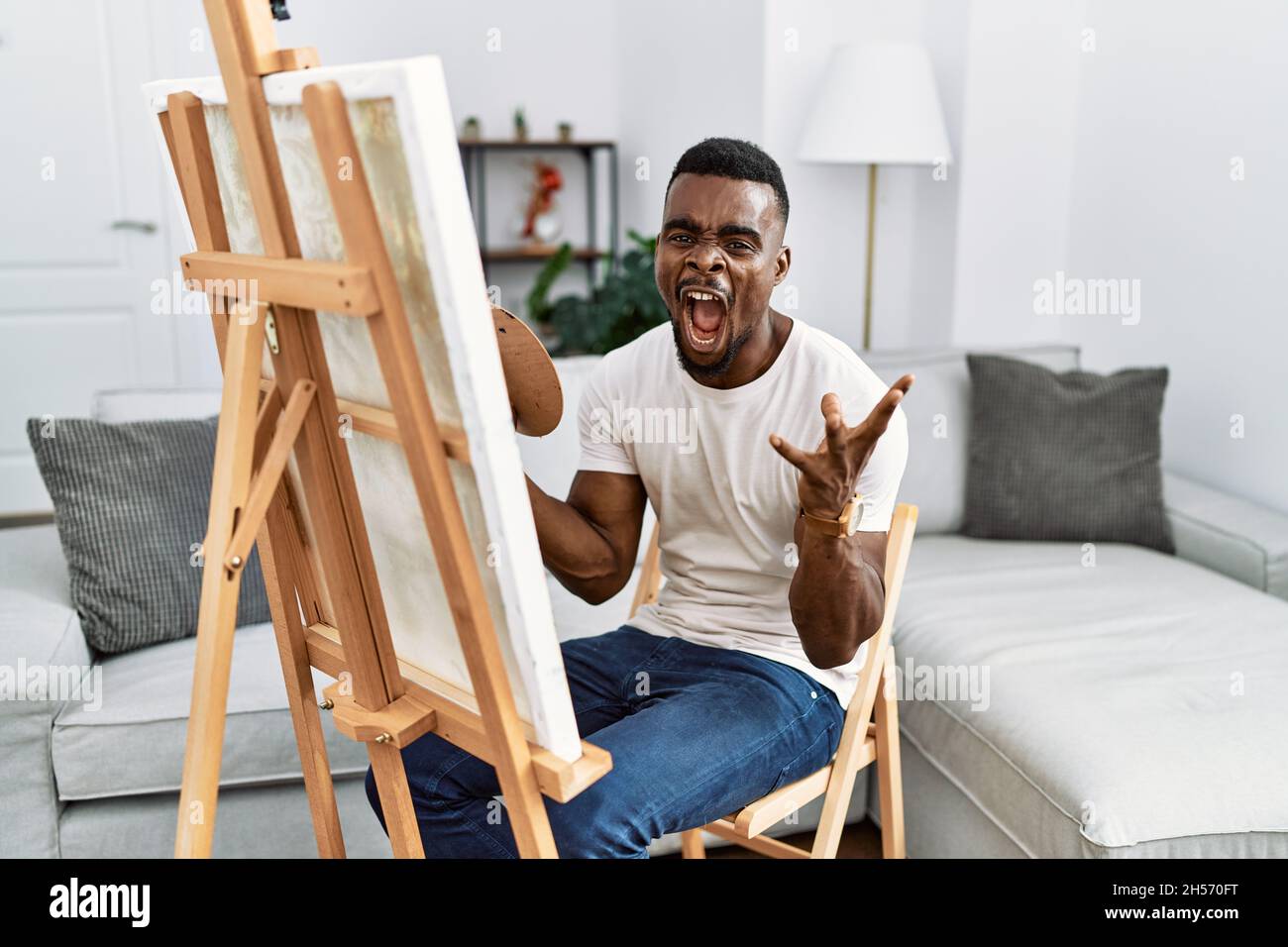 Young african man painting on canvas at home crazy and mad shouting and ...