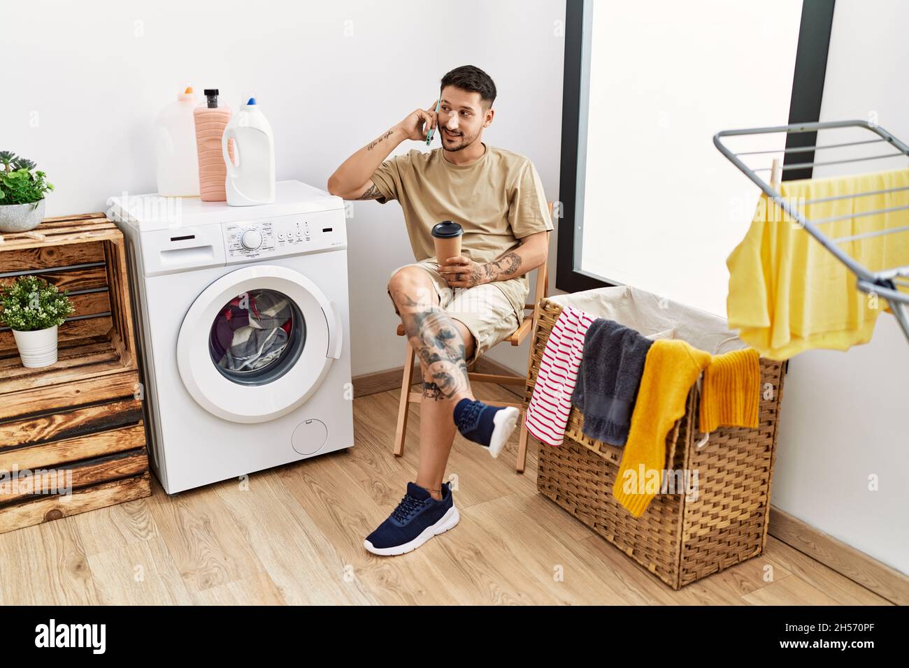 Young hispanic man waiting for washing machine speaking on the phone at ...