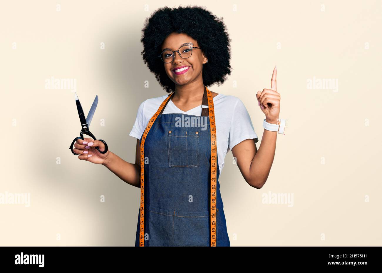 Young african american woman dressmaker designer wearing atelier apron holding scissors smiling ...