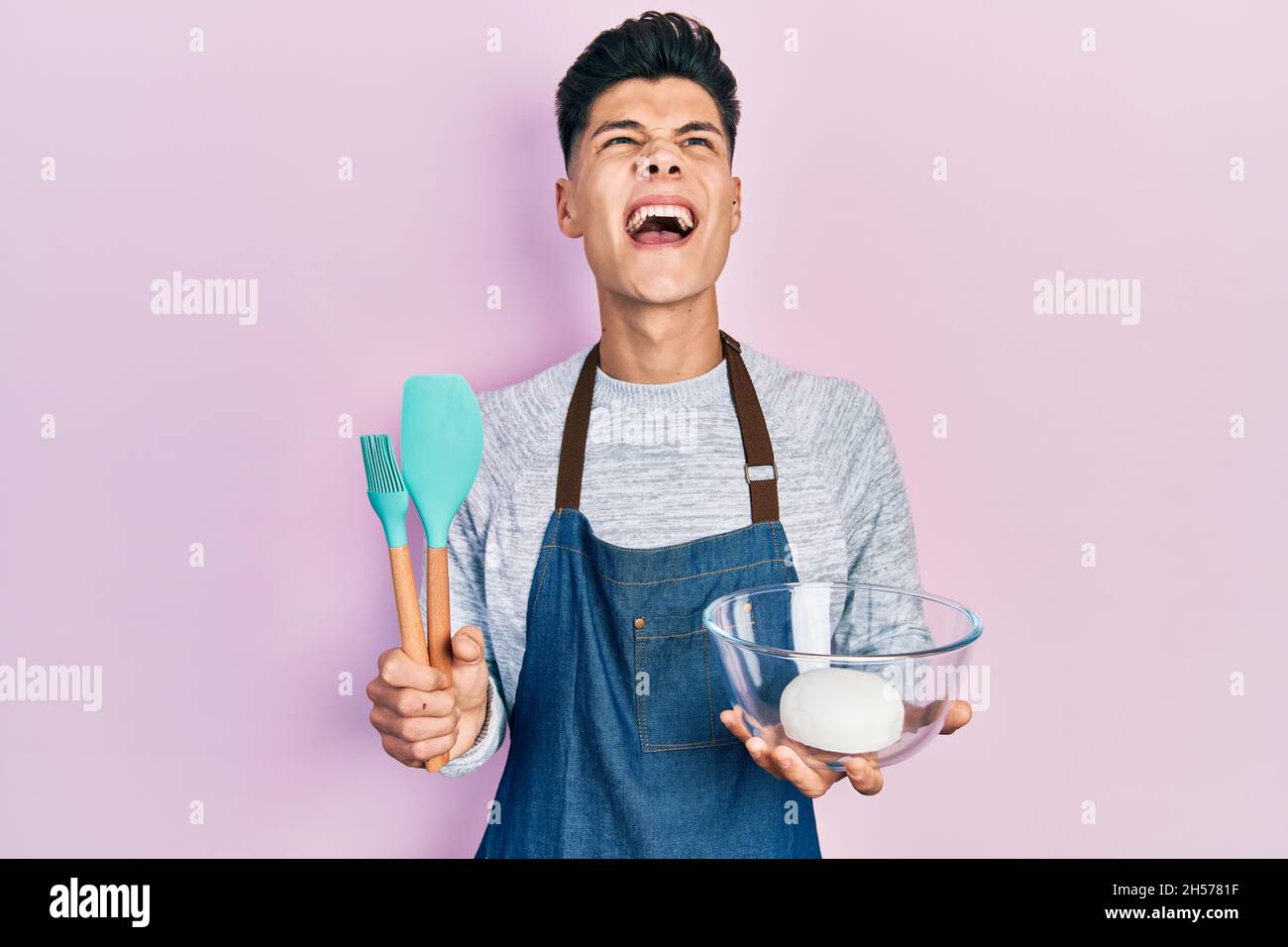 Young hispanic man holding bread dough and cooking tools angry and mad ...