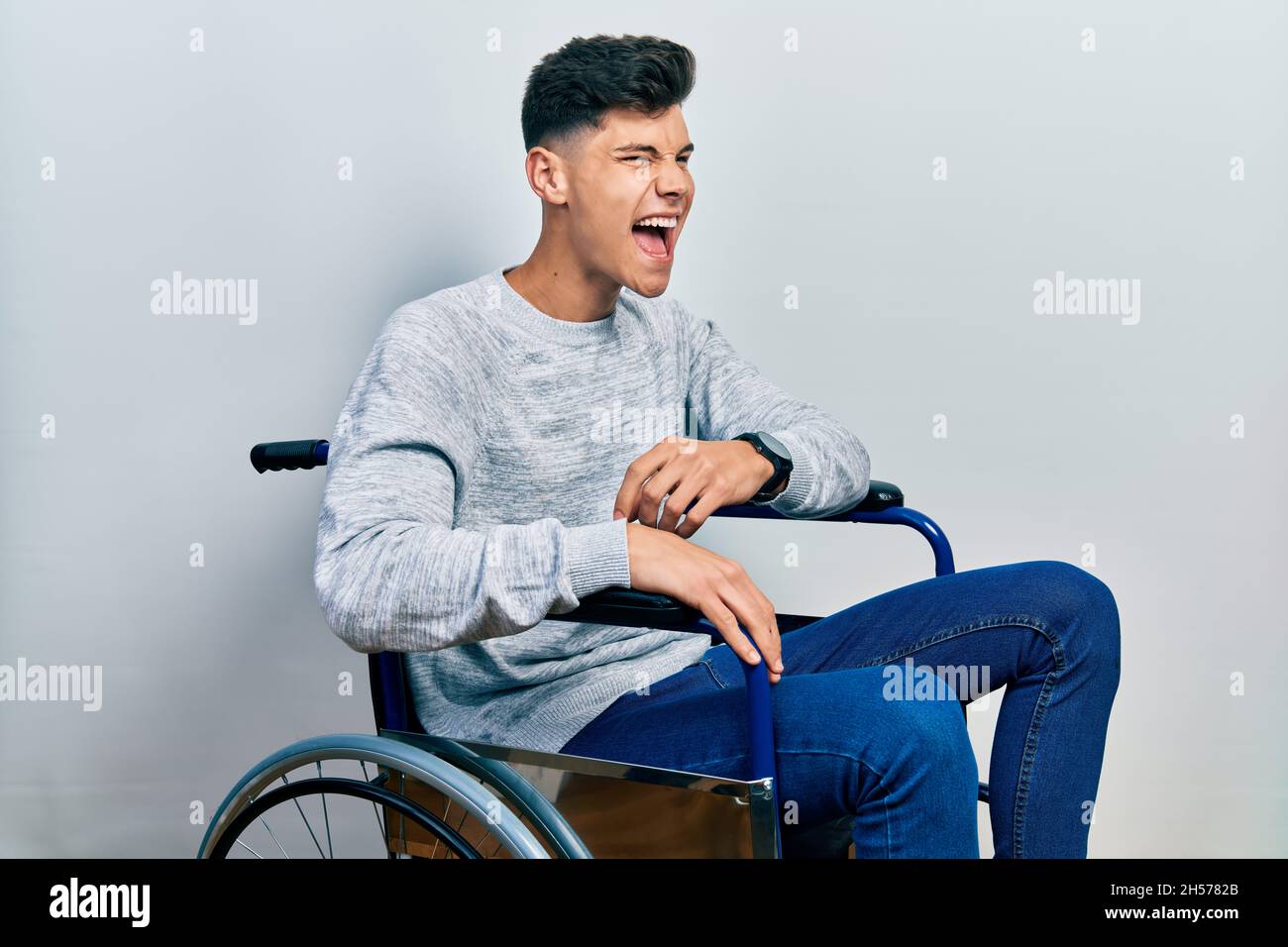 Young hispanic man sitting on wheelchair angry and mad screaming frustrated and furious ...