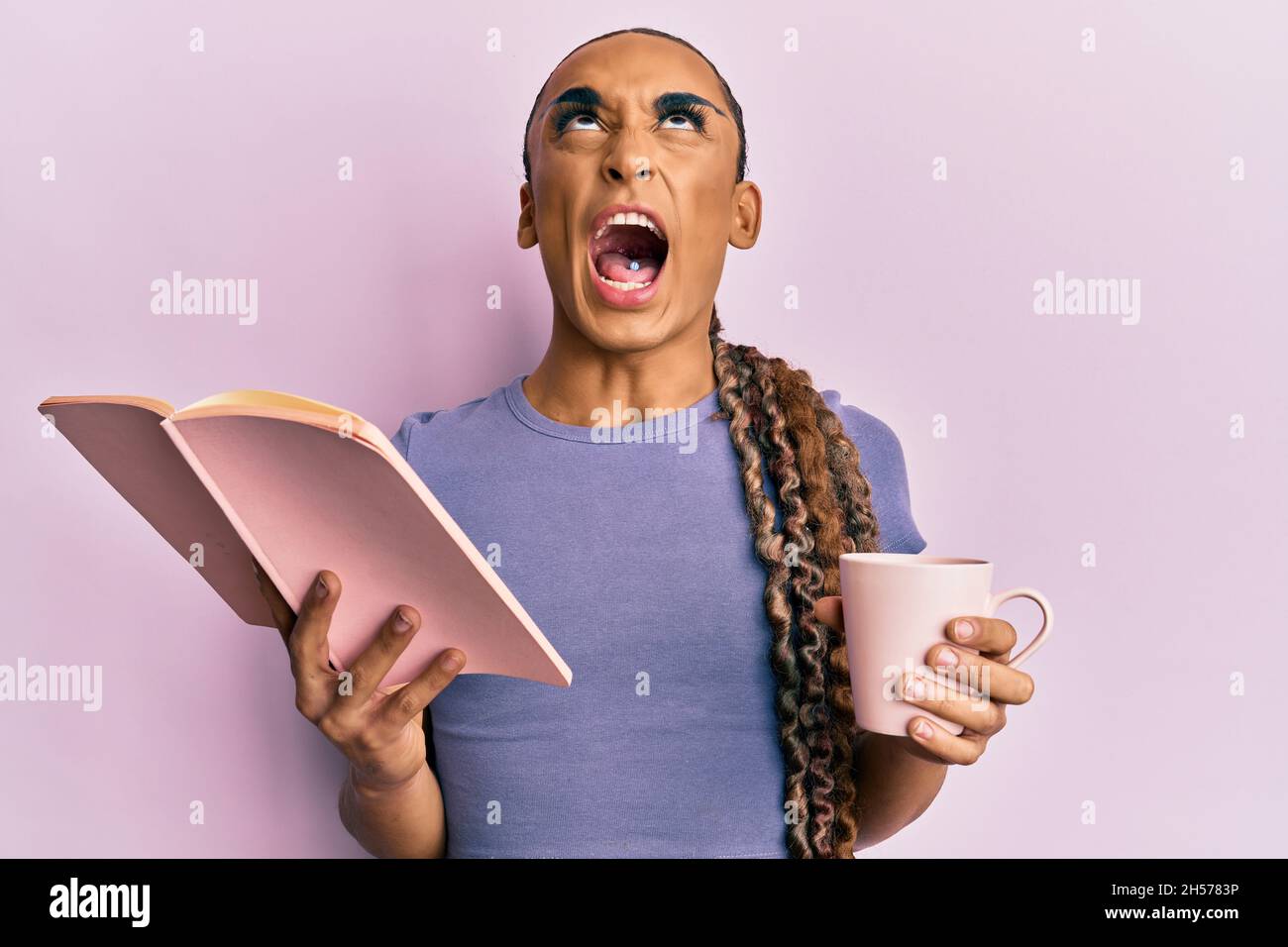 Hispanic man wearing make up and long hair reading book drinking cup of ...