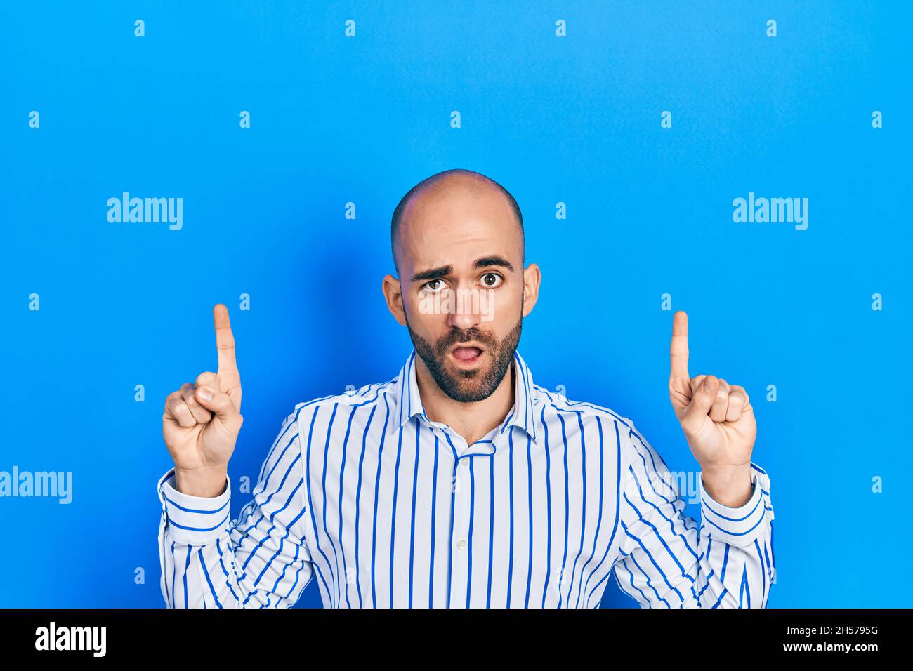 Young bald man pointing up with fingers in shock face, looking ...