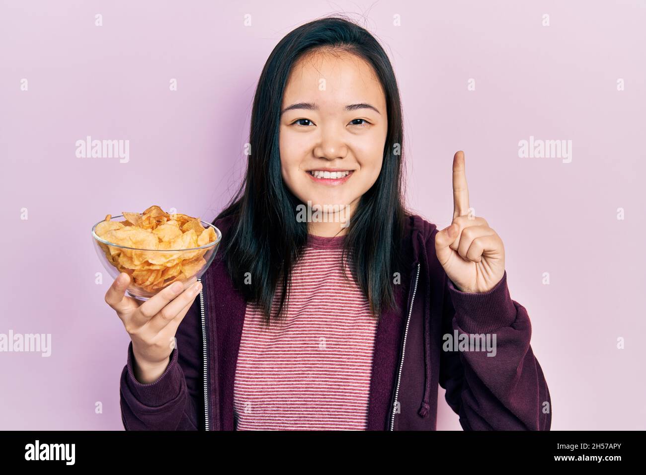 Young chinese girl holding potato chip smiling with an idea or question ...