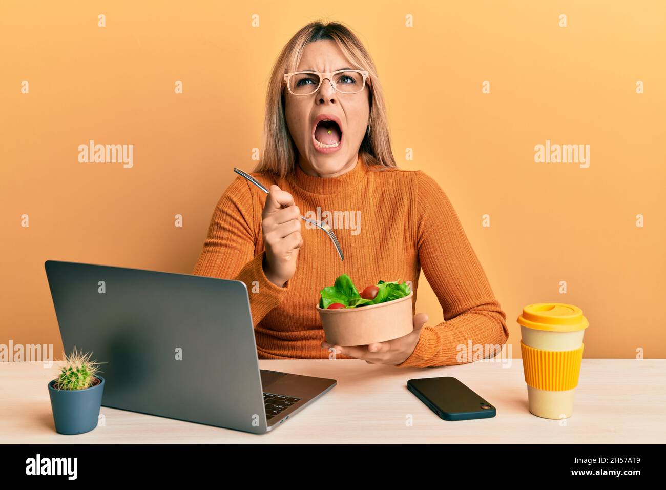 Young caucasian woman working at the office eating healthy salad angry ...
