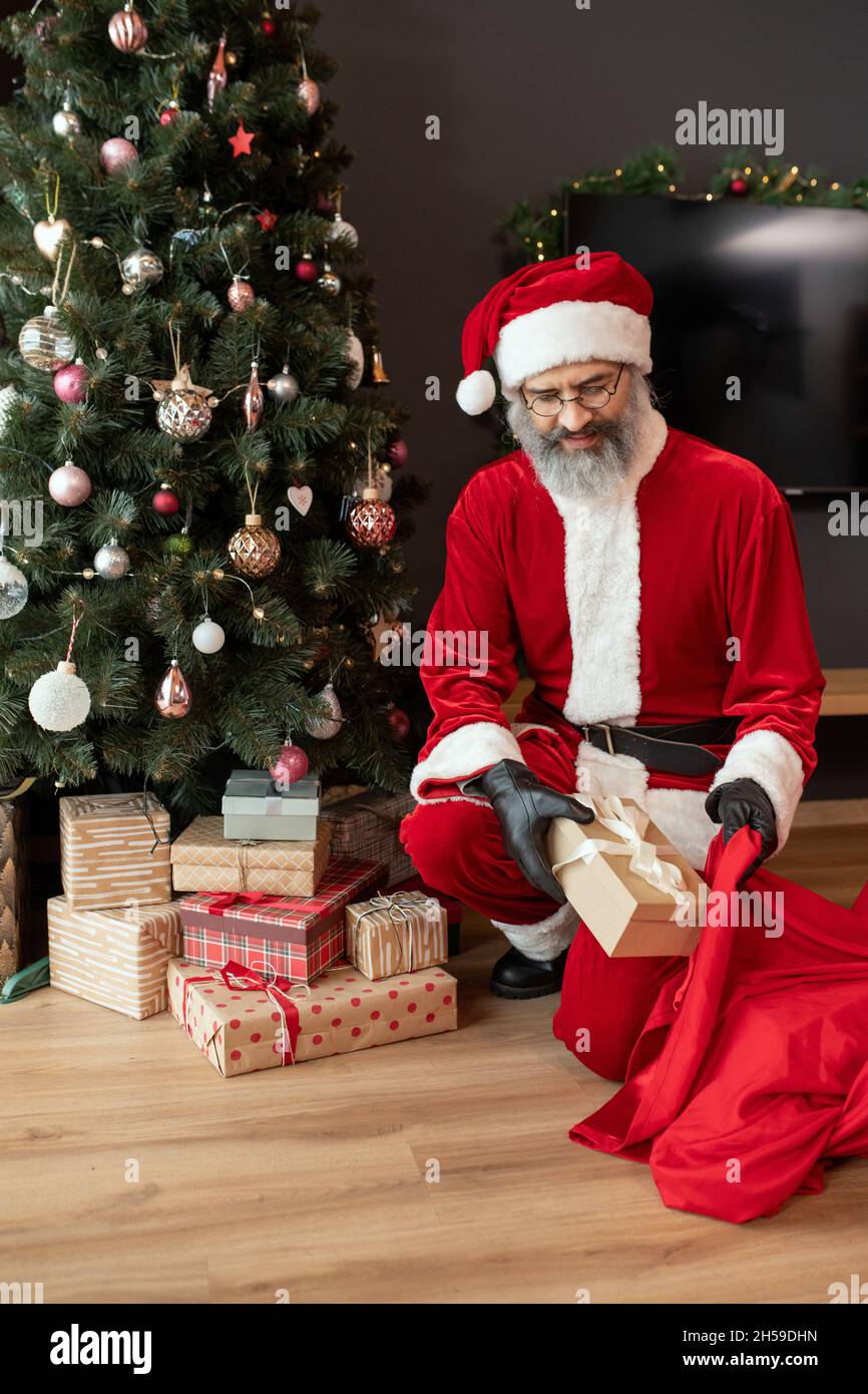 Smiling bad Santa in black gloves crouching at decorated tree and ...