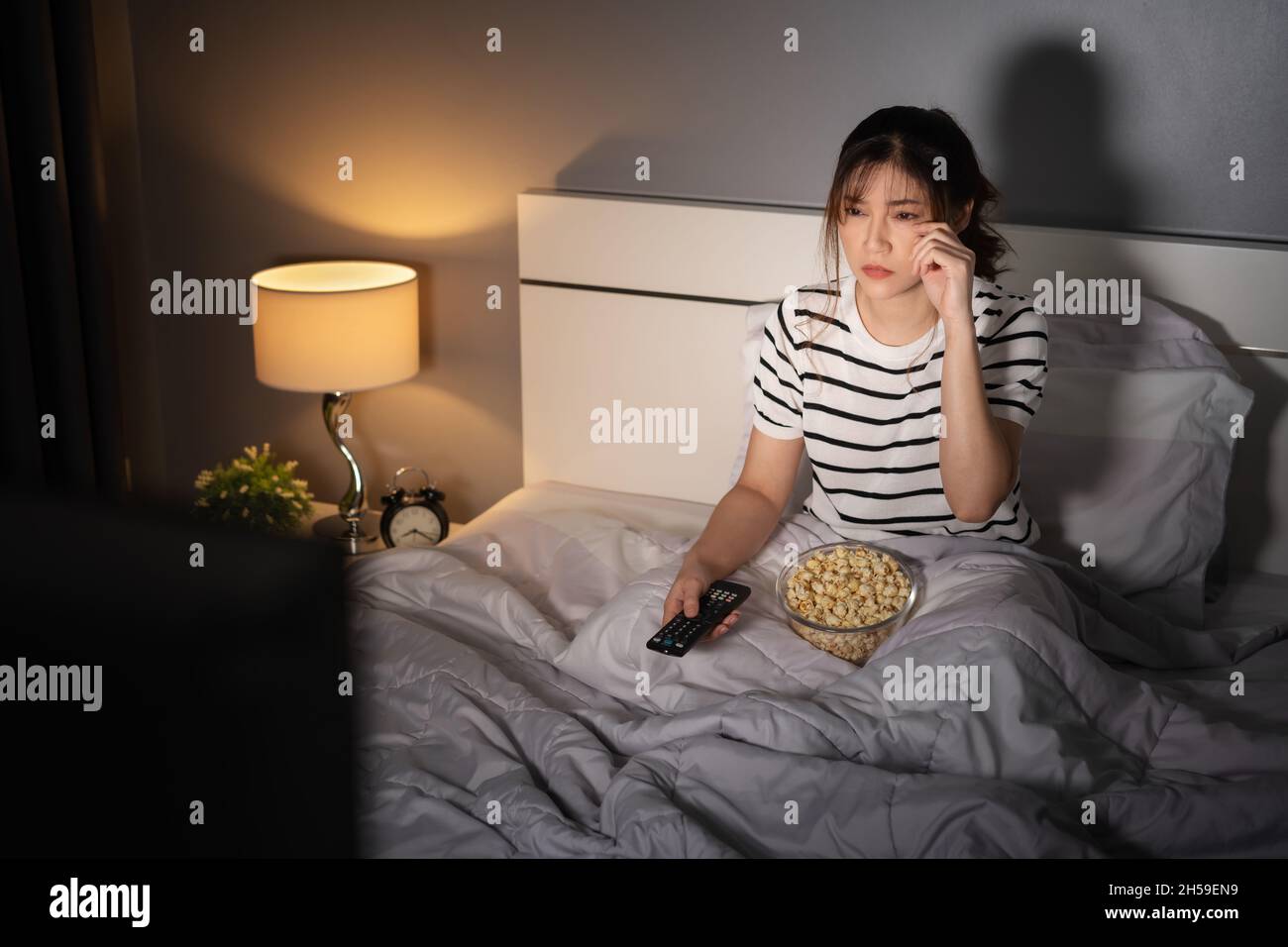 sad young woman watching television and crying on a bed at night ...
