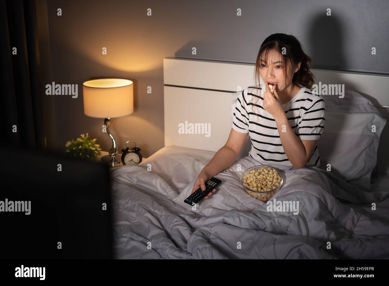 young woman watching TV and eating popcorn on a bed at night Stock ...