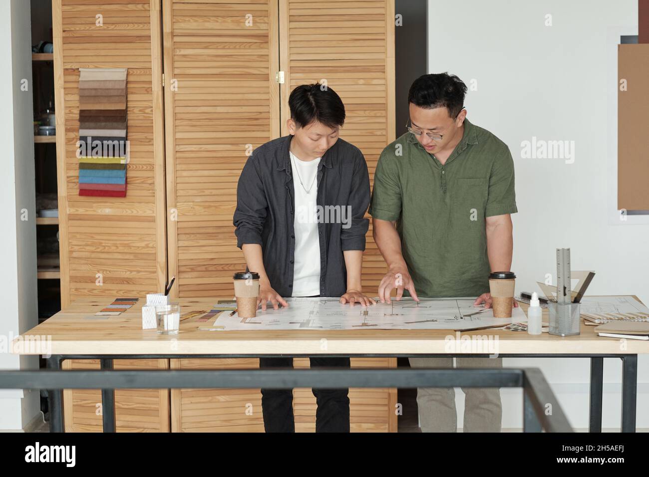 Two young contemporary engineers in casualwear standing by workplace ...