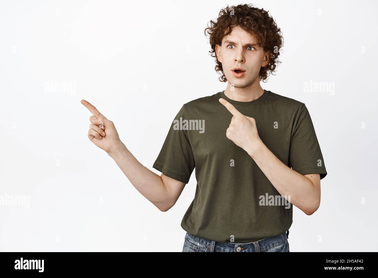 Shocked curly-haired man pointing fingers left, looking freak out and ...