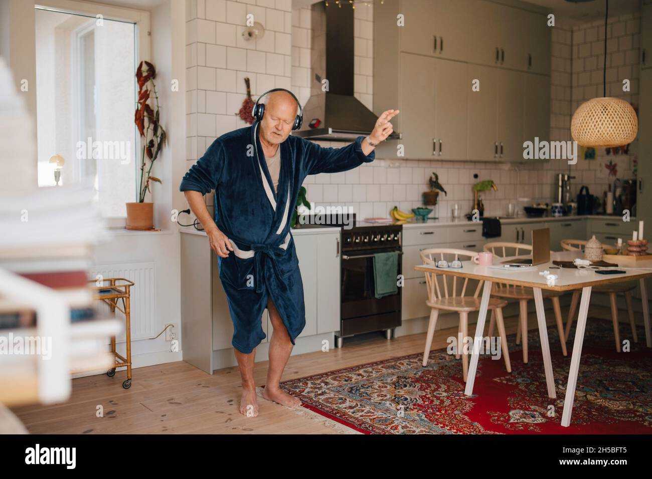 Senior man dancing while enjoying music through headphones in kitchen ...