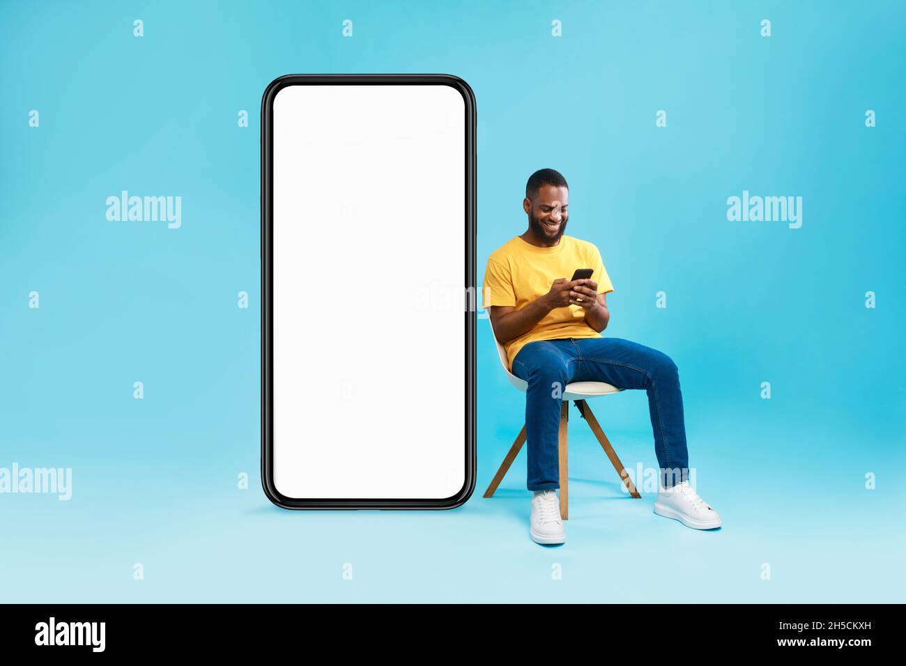 Cheerful young black man sitting on chair near big smartphone with ...