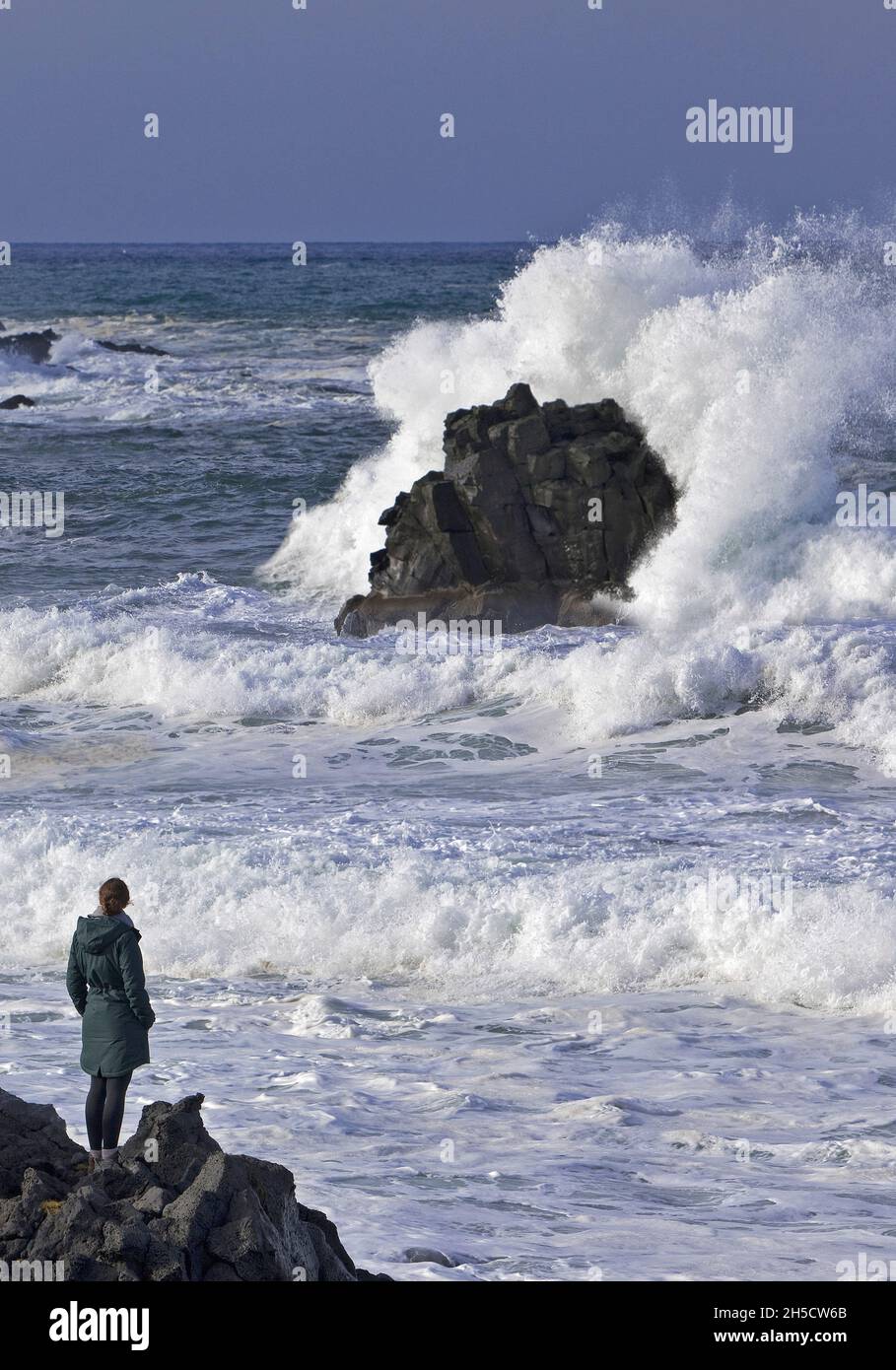 woman stands on the black lava coast and looking at the roaring ocean ...