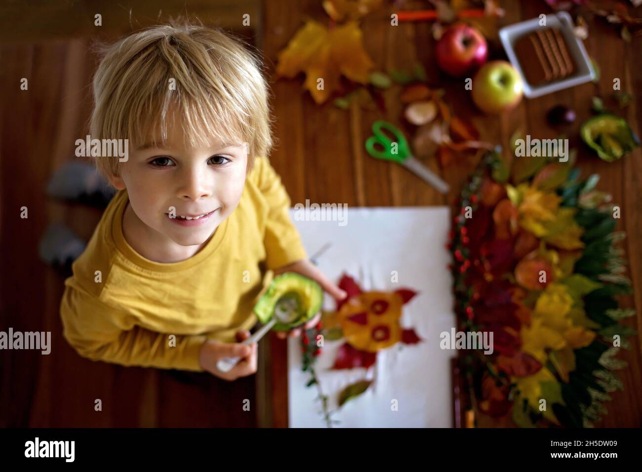 Sweet child, boy, eating avocado while applying leaves using glue while ...