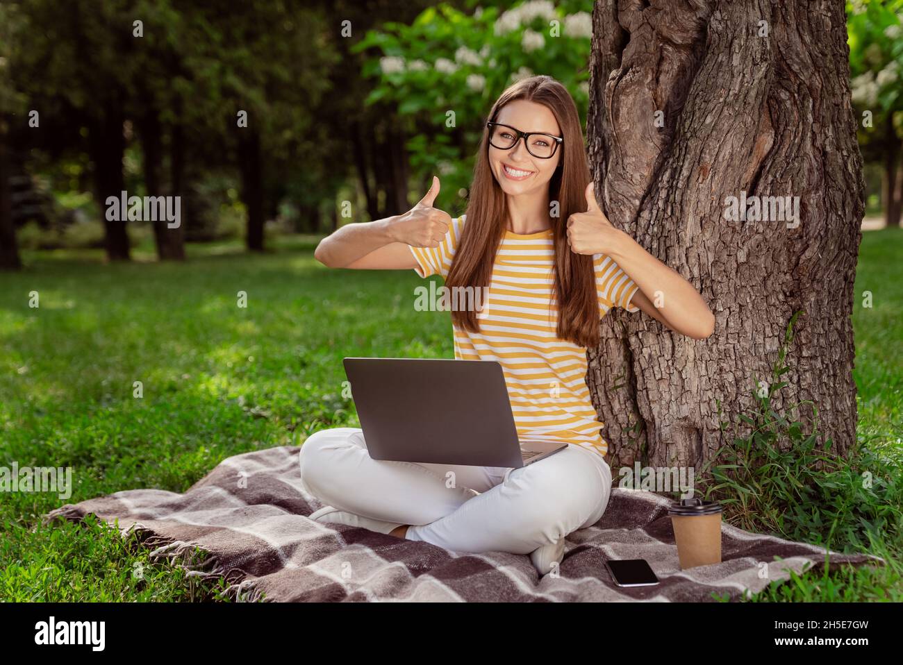 Portrait of attractive cheerful girl sitting on lawn using laptop showing double thumbup cool ...