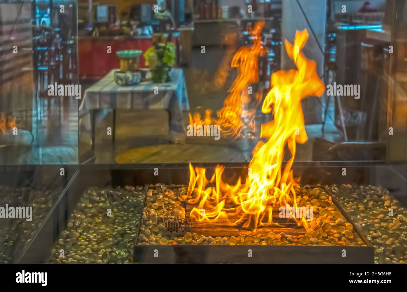 Dancing fire in a glass firebox with unrecognizable tables and chairs ...
