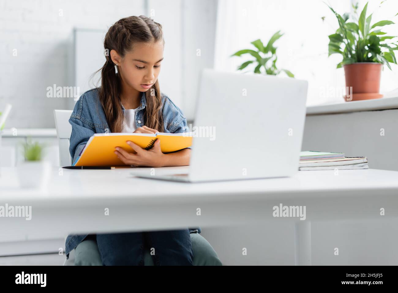 Preteen girl holding notebook near blurred laptop during online ...