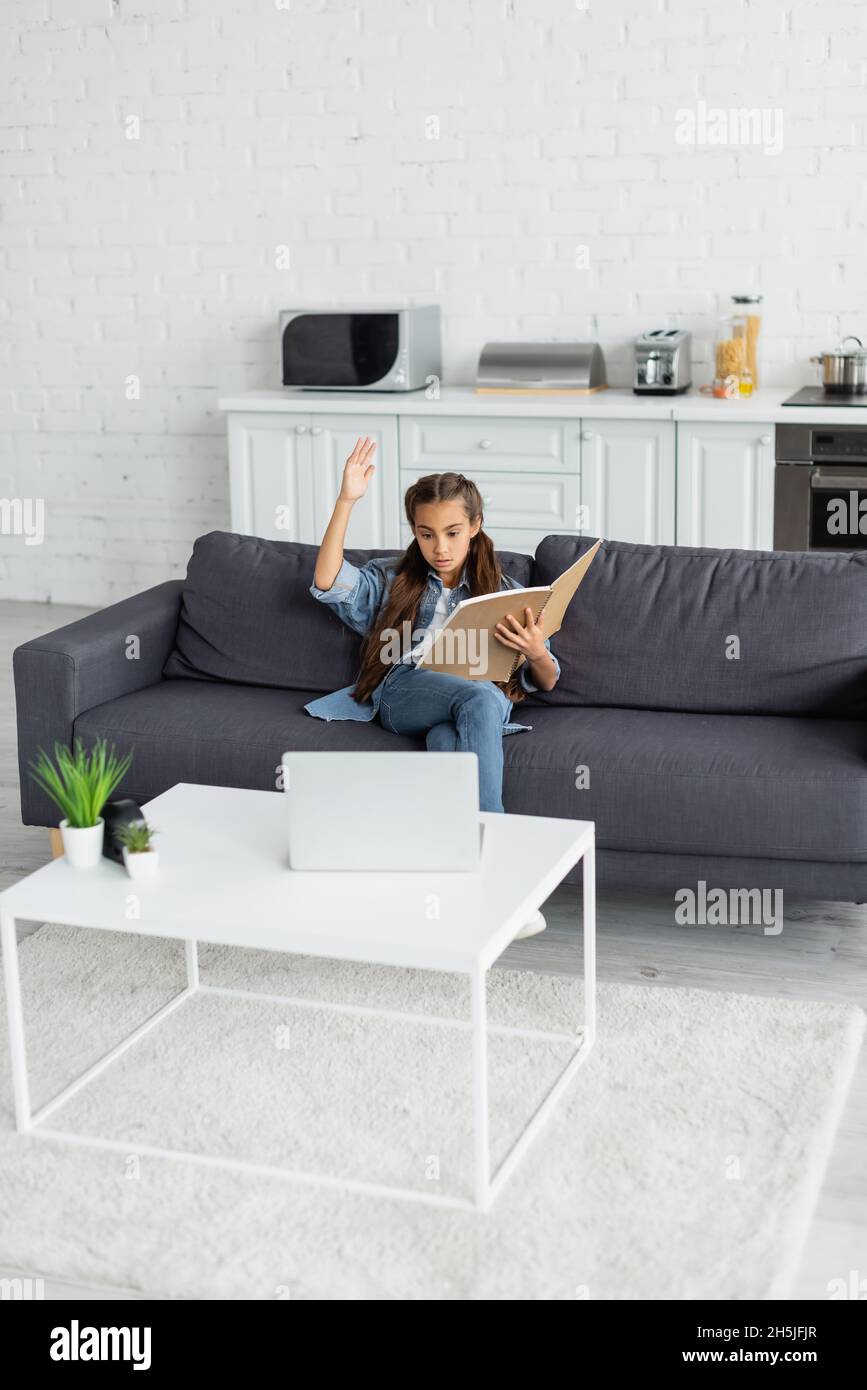 Preteen kid with notebook raising hand near laptop on couch at home ...