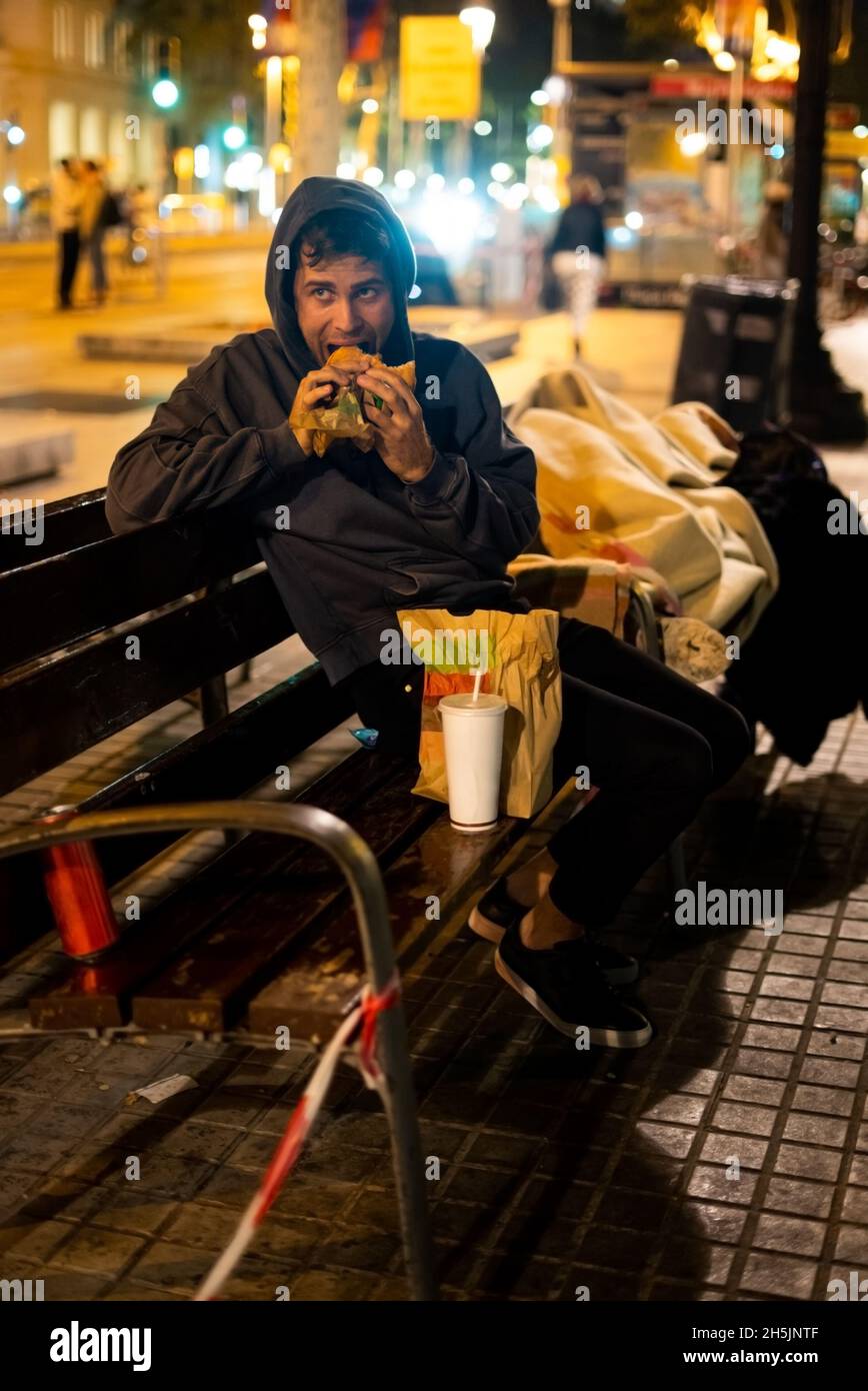 Hungry homeless man in hoodie looking away and biting burger while ...