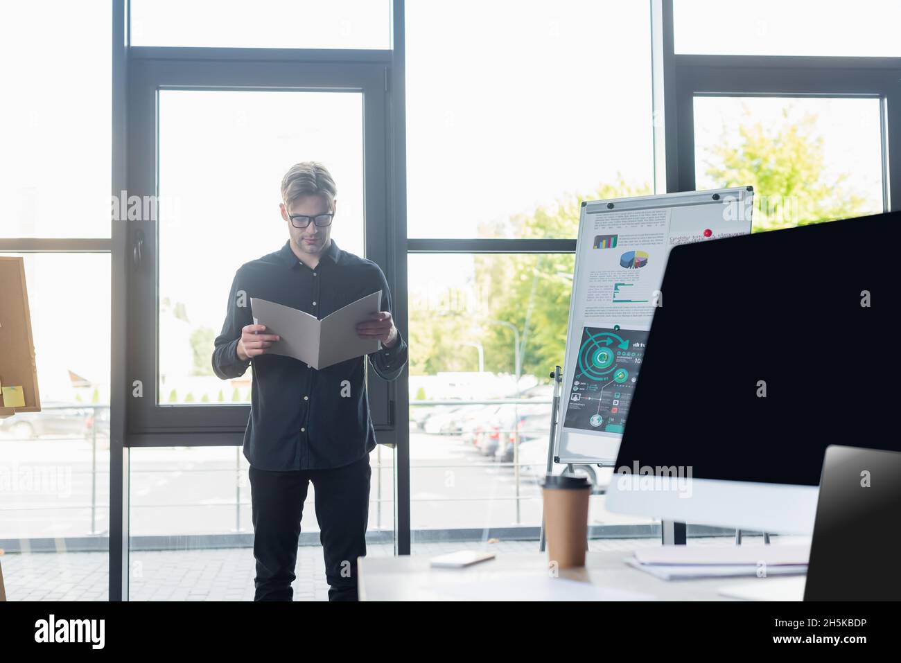 Programmer in eyeglasses holding paper folder near flip chart and devices in office Stock Photo ...