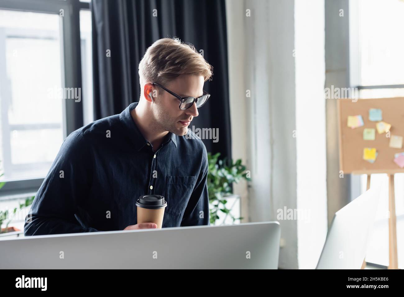Young programmer in earphone holding coffee near computer monitors ...