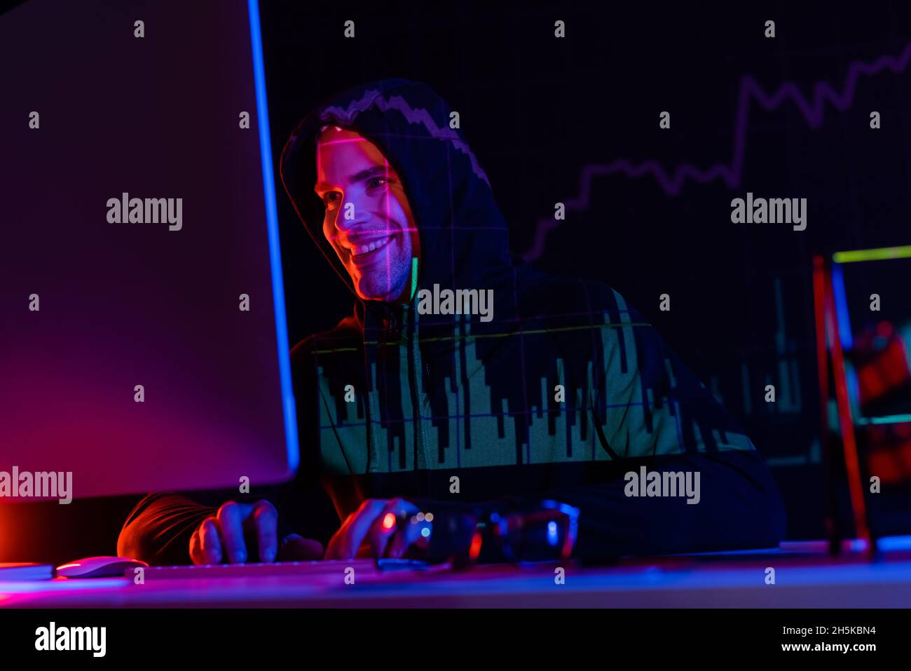 Smiling hacker using computer near eyeglasses and reflection of charts ...