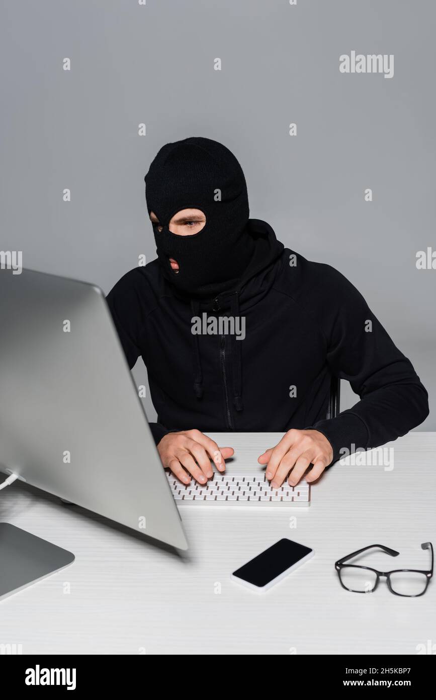 Hacker in balaclava using computer near smartphone and eyeglasses ...