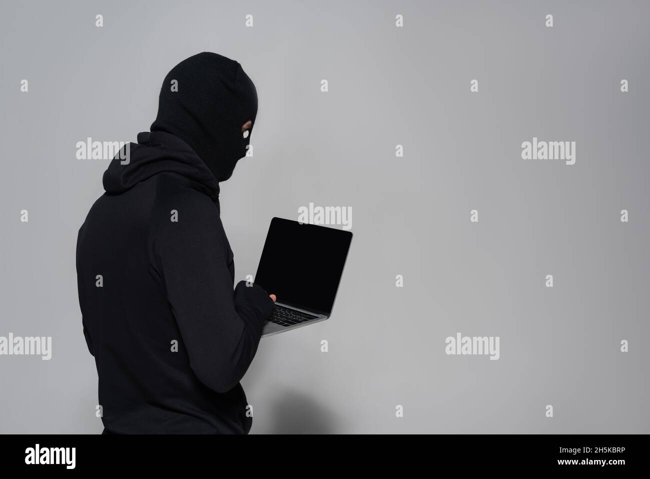 Hacker in balaclava using laptop with blank screen on grey background ...