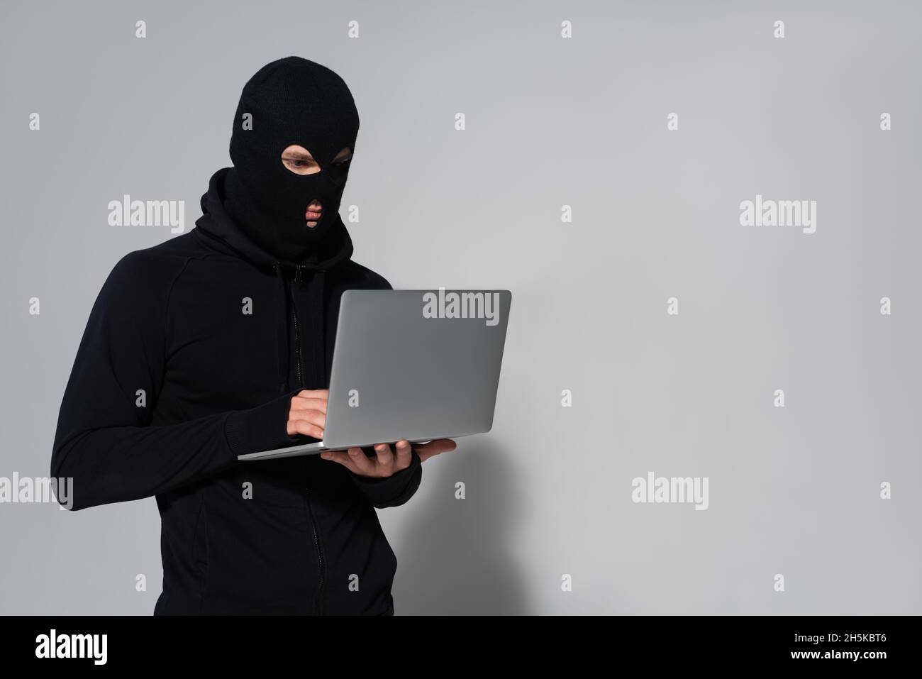 Hacker in balaclava using laptop on grey background Stock Photo - Alamy