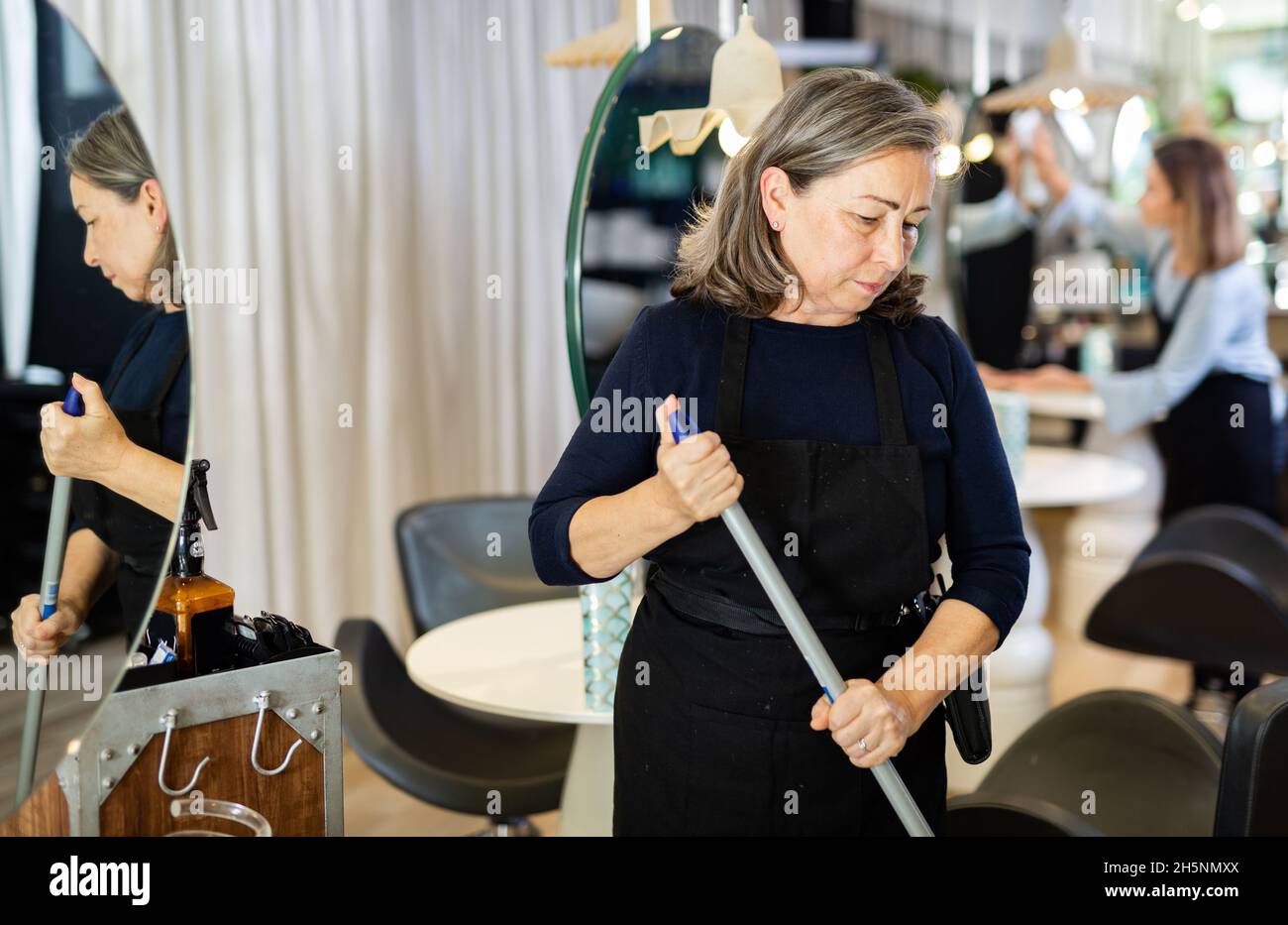 Elderly female hairdresser sweeping floor in modern barbershop Stock ...