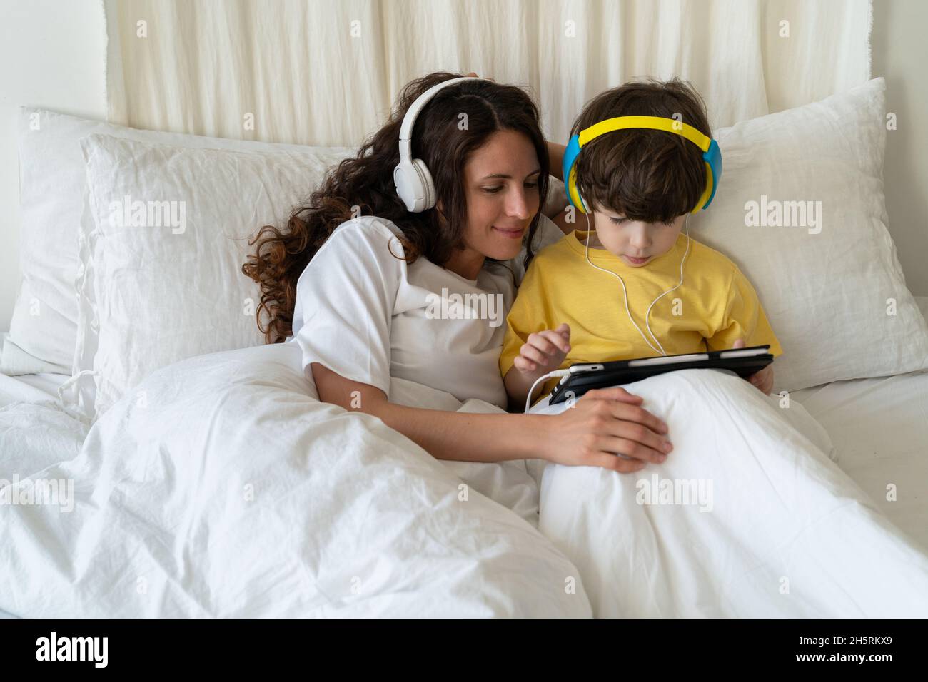 Family morning in bedroom: mom in headphones look at little kid son ...