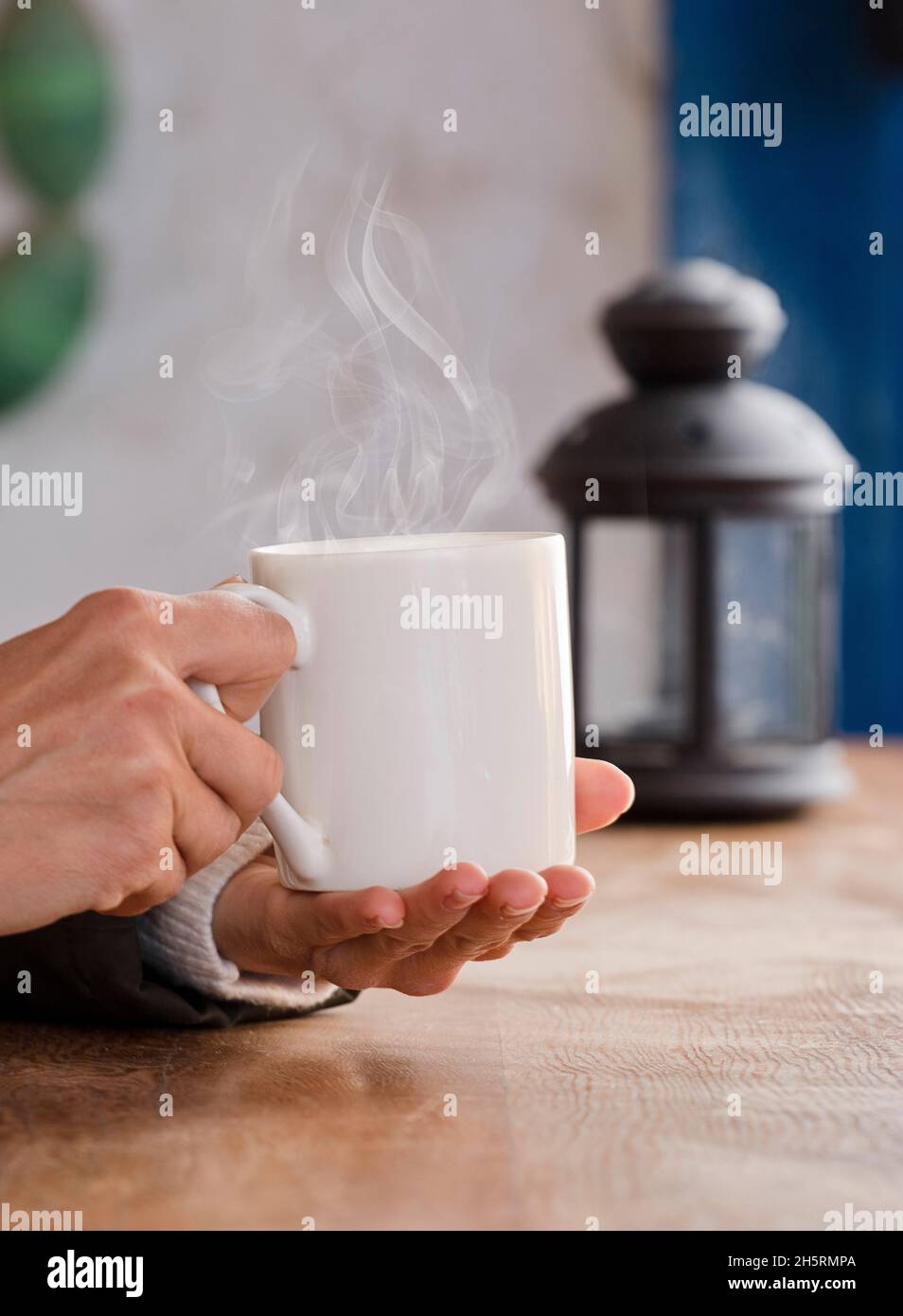 white cup with hot drink and smoke, hands warming up from the heat, at home. Focus on the cup ...