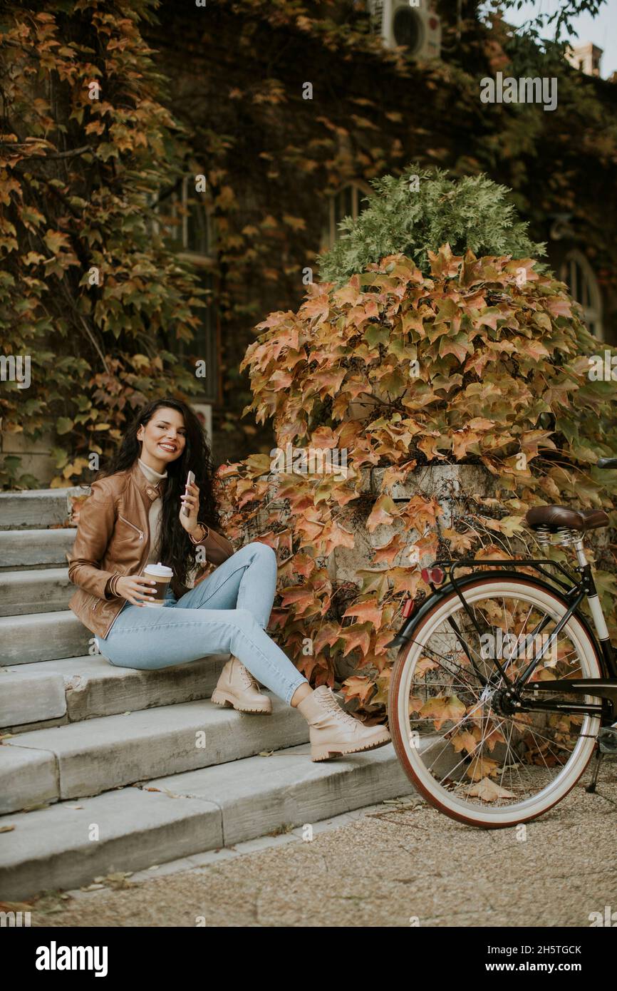 Pretty young woman with mobile phone drink coffee to go at the stairs by the bicycle on autumn ...