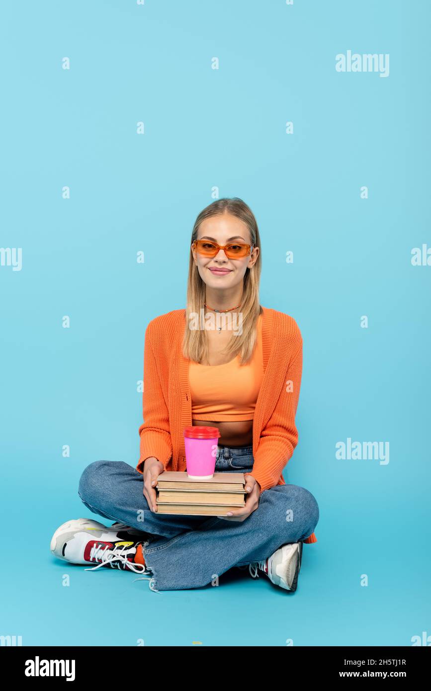 Smiling student in sunglasses holding books and paper cup while sitting ...