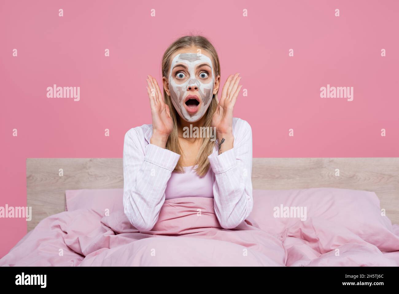 Shocked woman with clay mask on face looking at camera on bed isolated ...