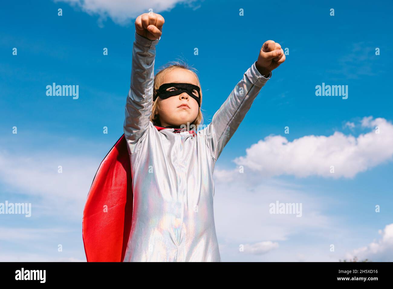 From below small girl in superhero costume raising outstretched fists ...