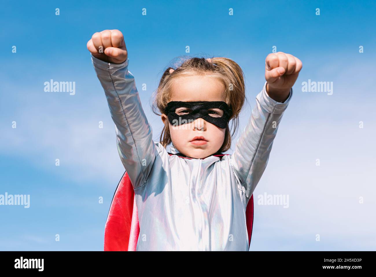 From below small girl in superhero costume raising outstretched fists ...