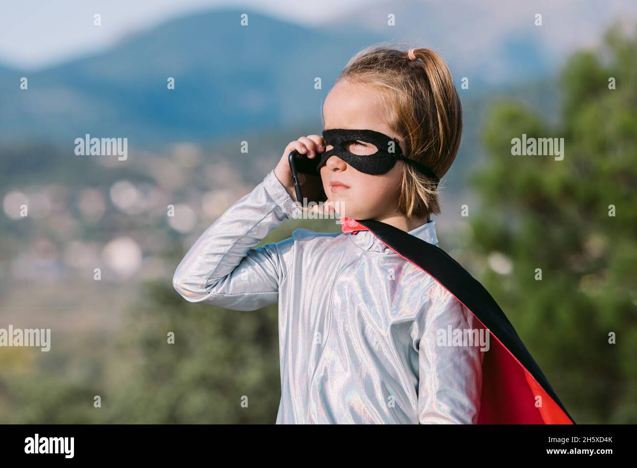 Self assured girl in eye mask superhero costume with cape having phone ...