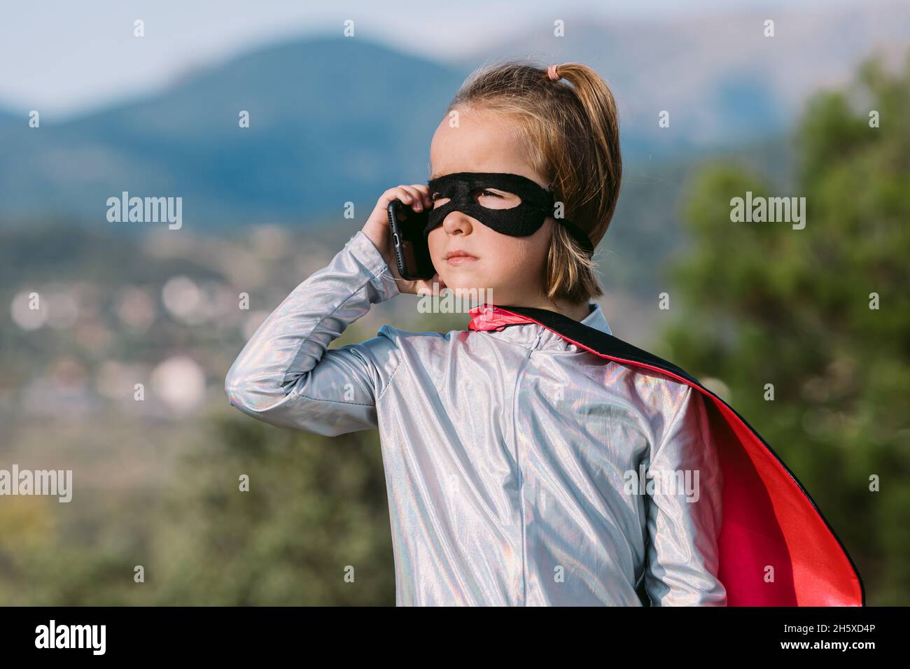 Self assured girl in eye mask superhero costume with cape having phone ...