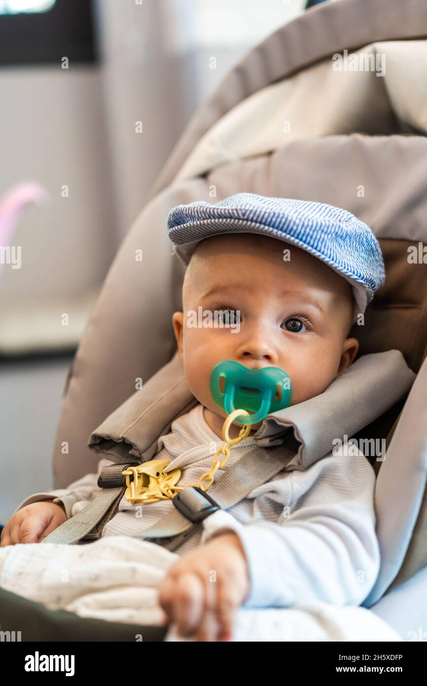 Adorable baby in hat looking away and sucking pacifier while sitting in ...