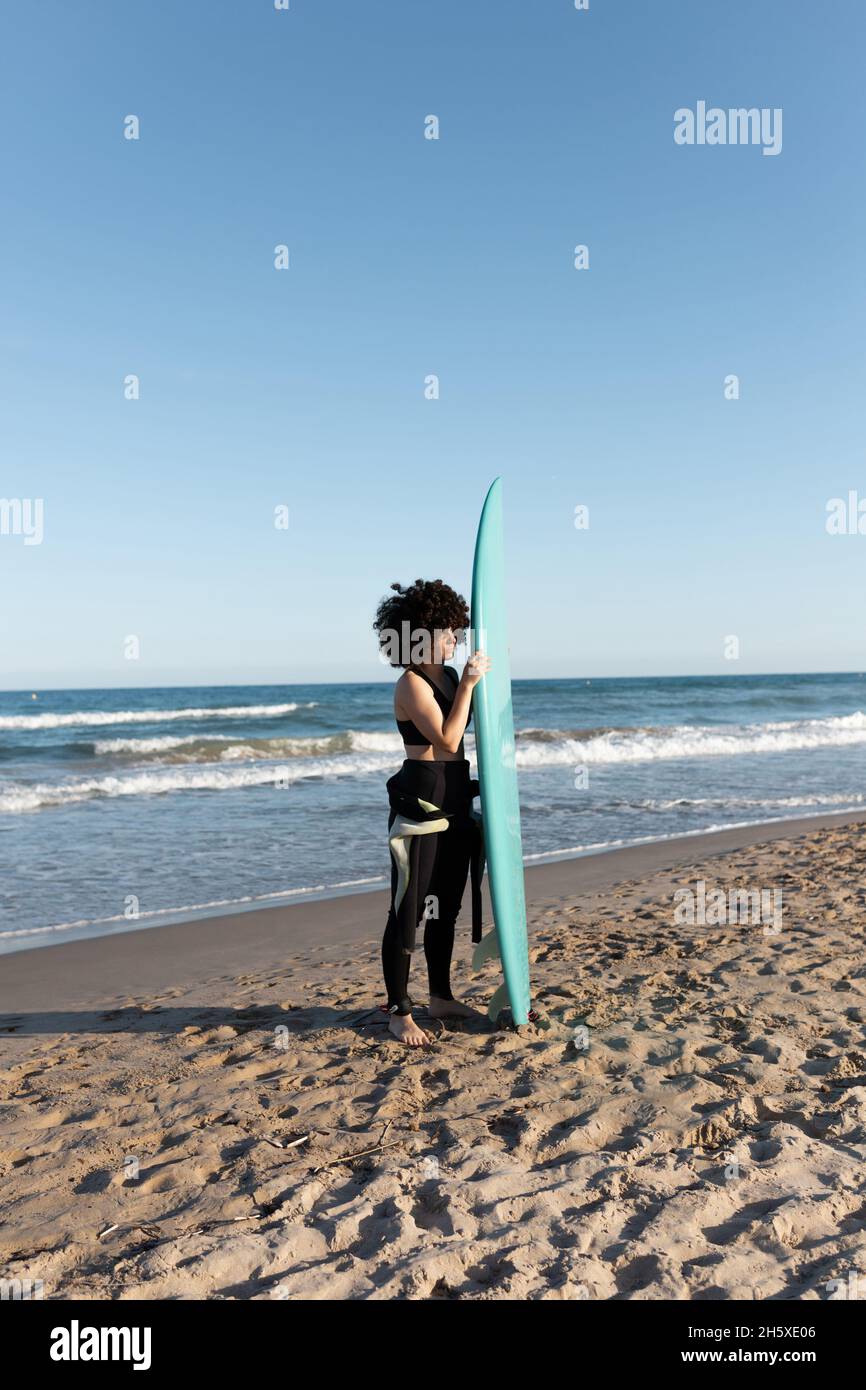 Side view of young female surfer in wetsuit with surfboard standing ...