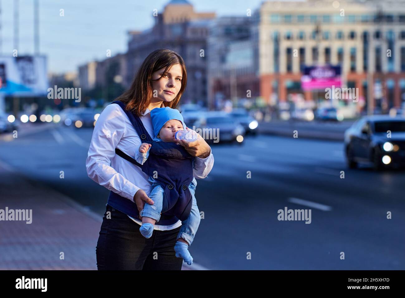 Woman feeds baby in sling from baby bottle in front of city street with ...