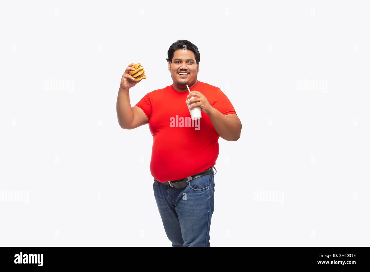 Portrait of a fat man holding a drink and burger in hand against plain ...