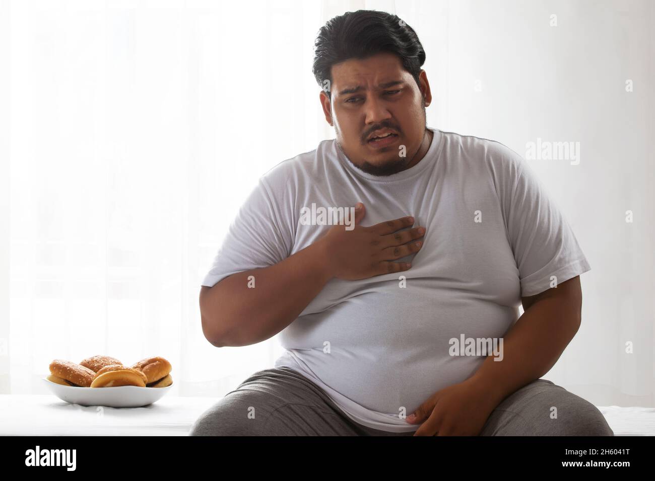 A fat man sitting,keeping hand on his chest due to pain with a plate of ...